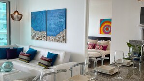 Uma cozinha moderna com bancadas de granito, armários brancos e uma ilha central. Há duas luminárias pendentes, uma janela grande e uma pintura abstrata colorida na parede.