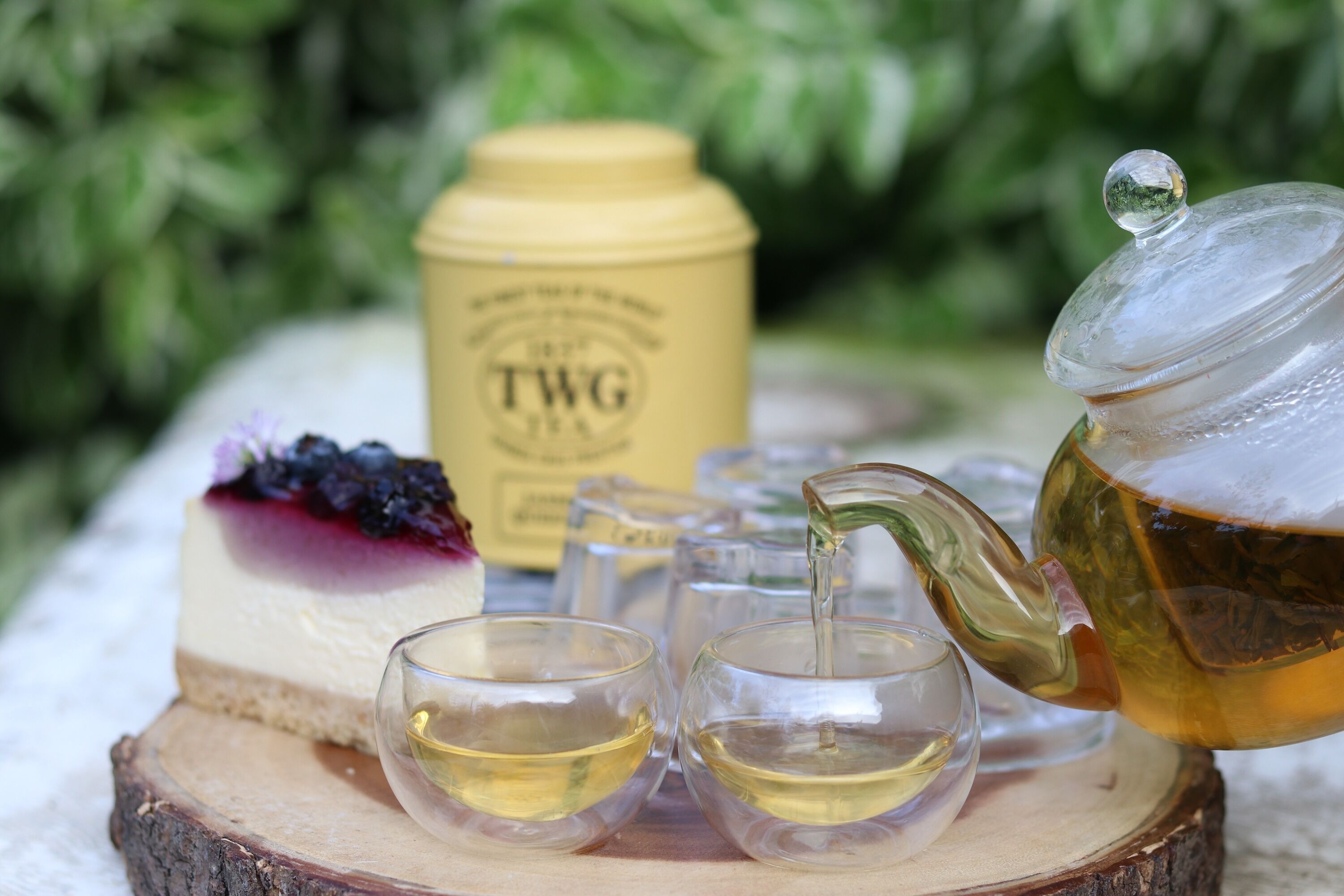 Uma mesa de madeira com uma chaleira de vidro a servir chá em duas taças, um frasco com a inscrição "TWG" e uma sobremesa com cobertura roxa.