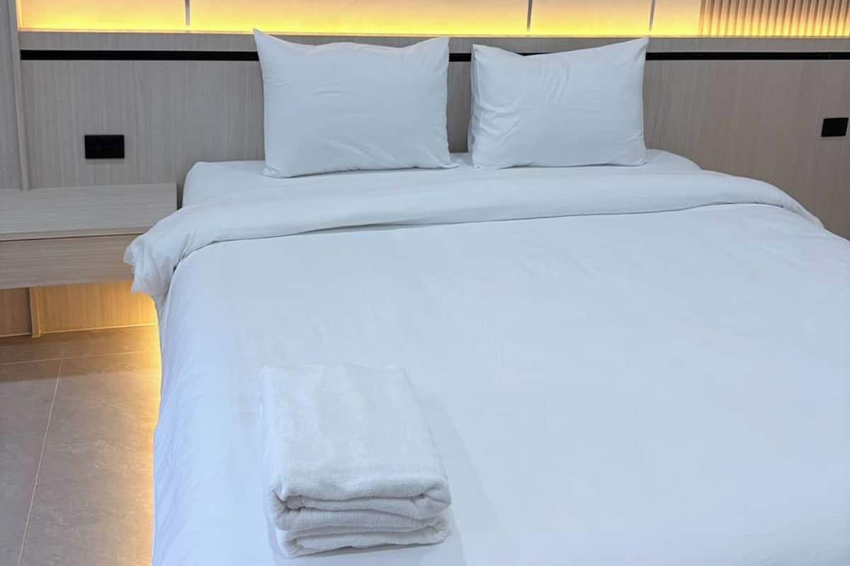 Cama bem arrumada com lençóis e almofadas brancos, uma mesa de cabeceira e uma parede com painéis verticais e luz quente.