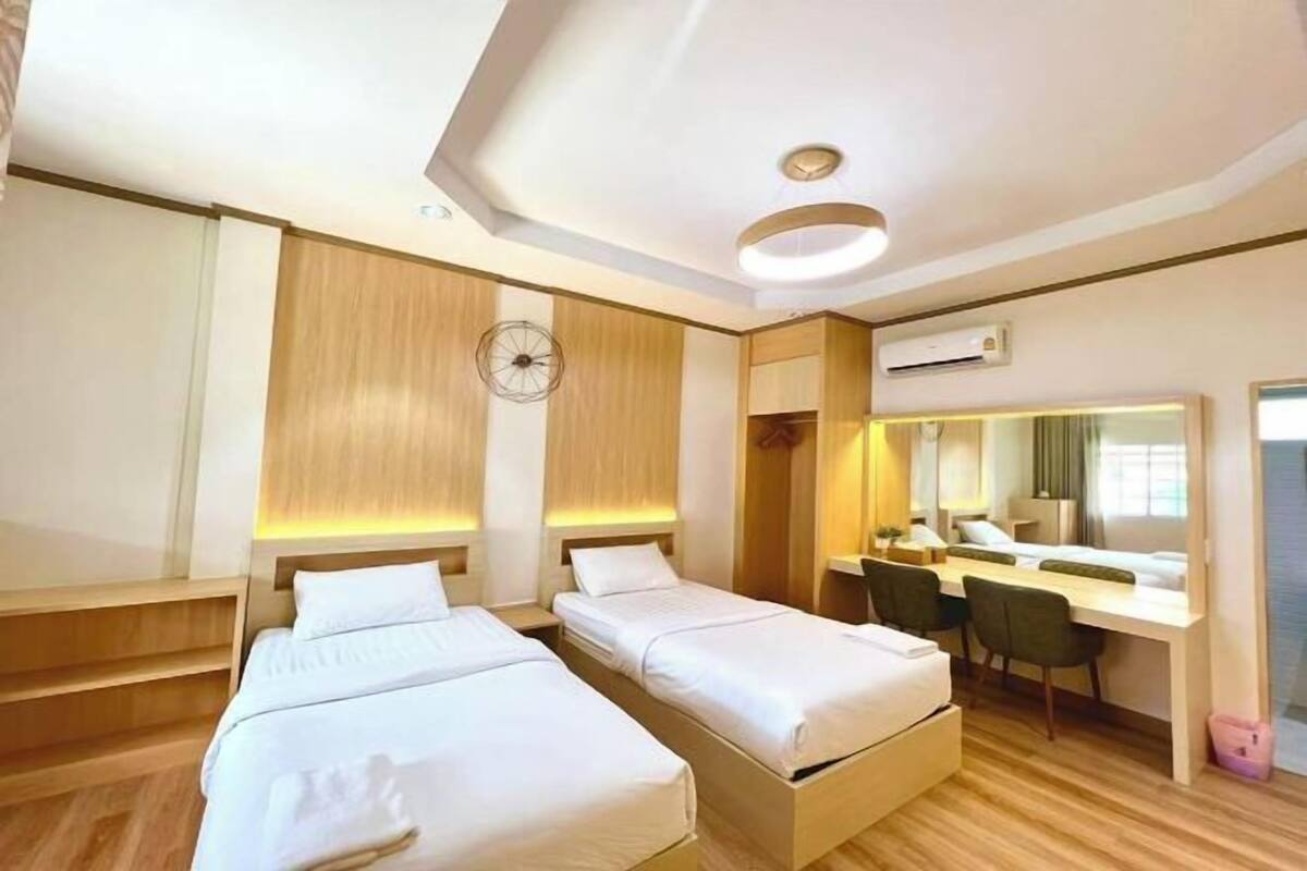 Quarto de hotel com duas camas, uma escrivaninha, uma cadeira, um espelho e um relógio.