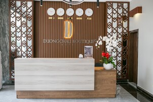 Balcão de receção com acabamento em madeira e uma bancada de mármore clara. Em cima do balcão, existem relógios com diferentes fusos horários indicados, e um grande letreiro iluminado com a letra 'D'.