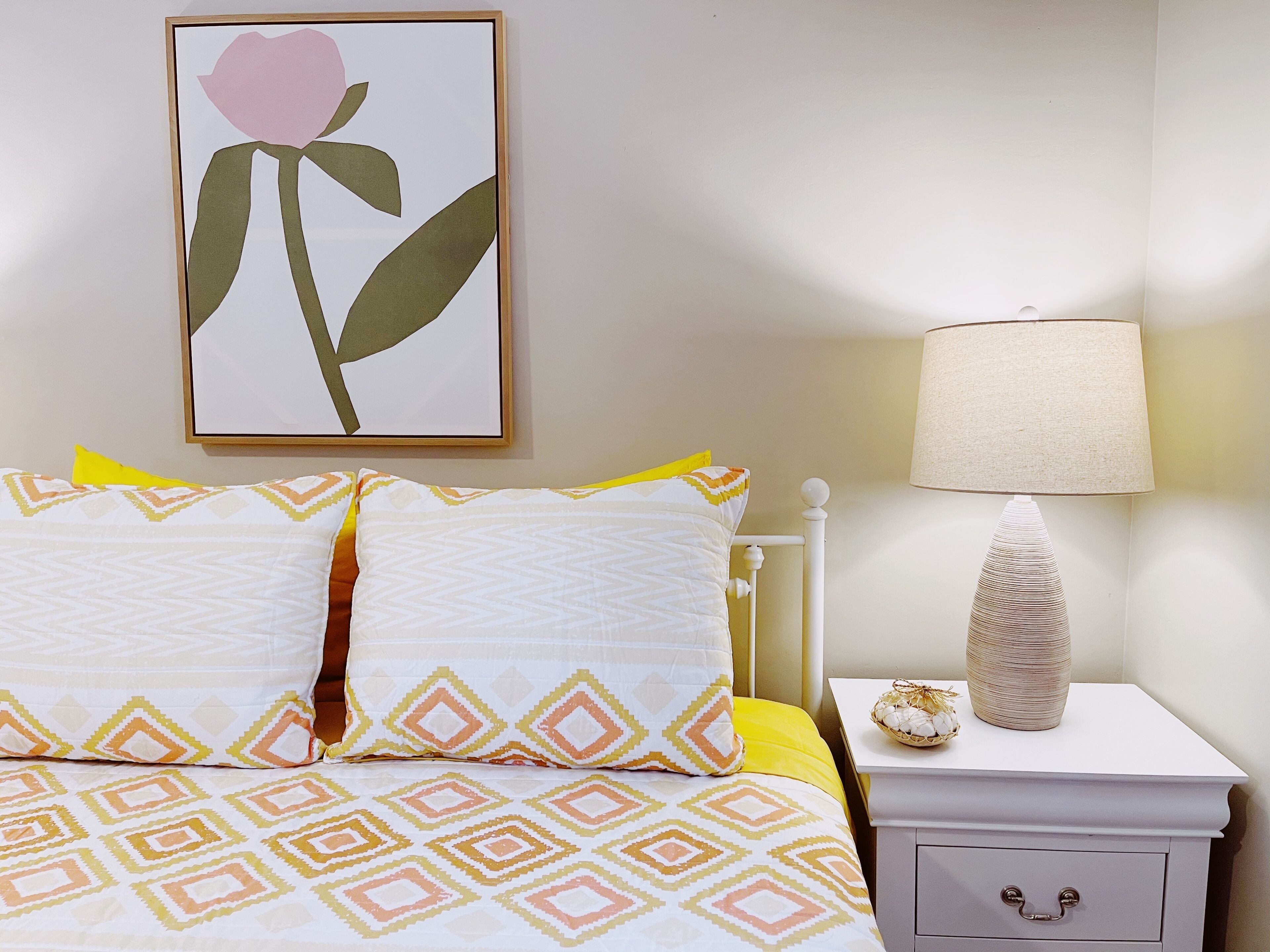 Cama bem arrumada com roupa de cama estampada, mesa de cabeceira com abajur e uma obra de arte emoldurada na parede.