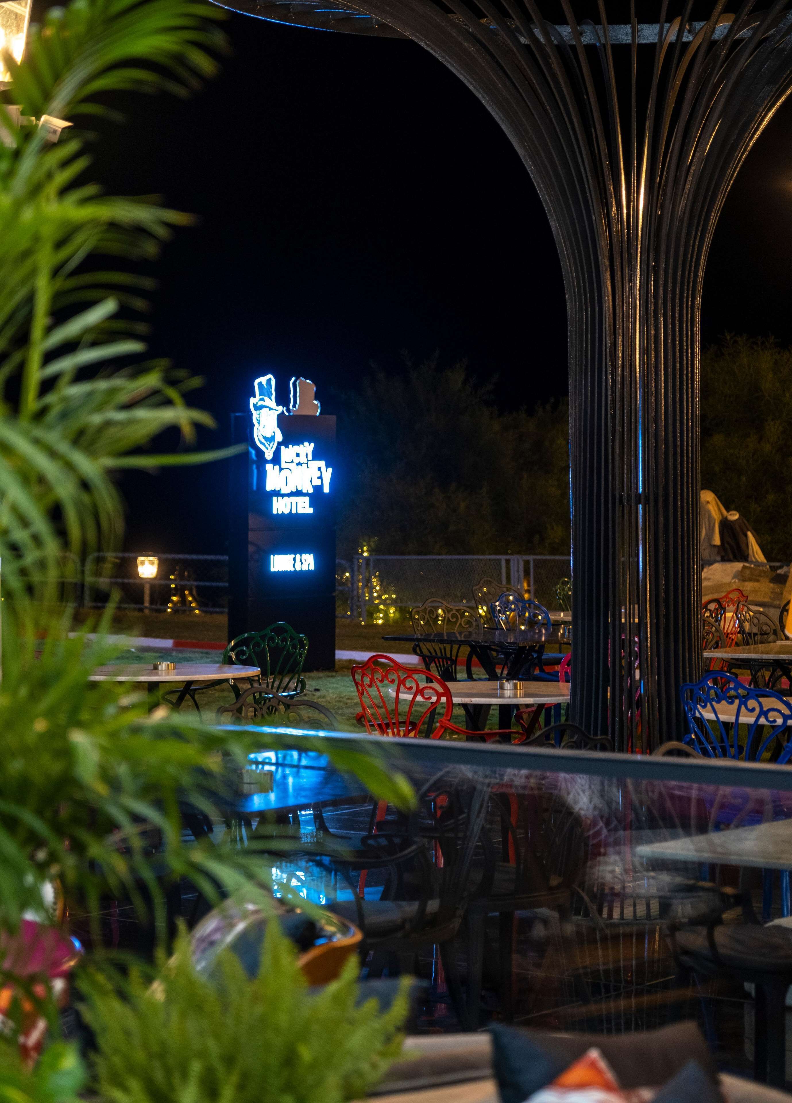 Vista noturna de um restaurante com mesas ao ar livre, um letreiro de neon que diz 'Monkey Hotel' e uma estrutura de metal decorativa.