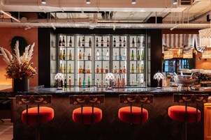 Uma área de bar com exposição de vinhos e bebidas, banquetas acolchoadas vermelhas e uma planta decorativa.