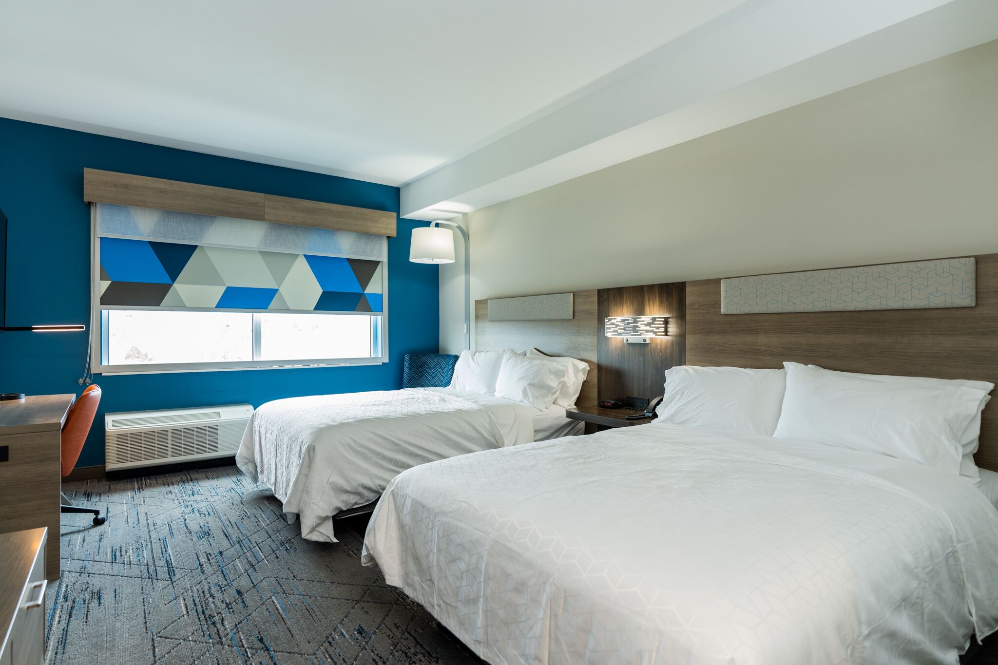 Quarto de hotel com duas camas, uma escrivaninha e uma janela com persiana de padrão geométrico.