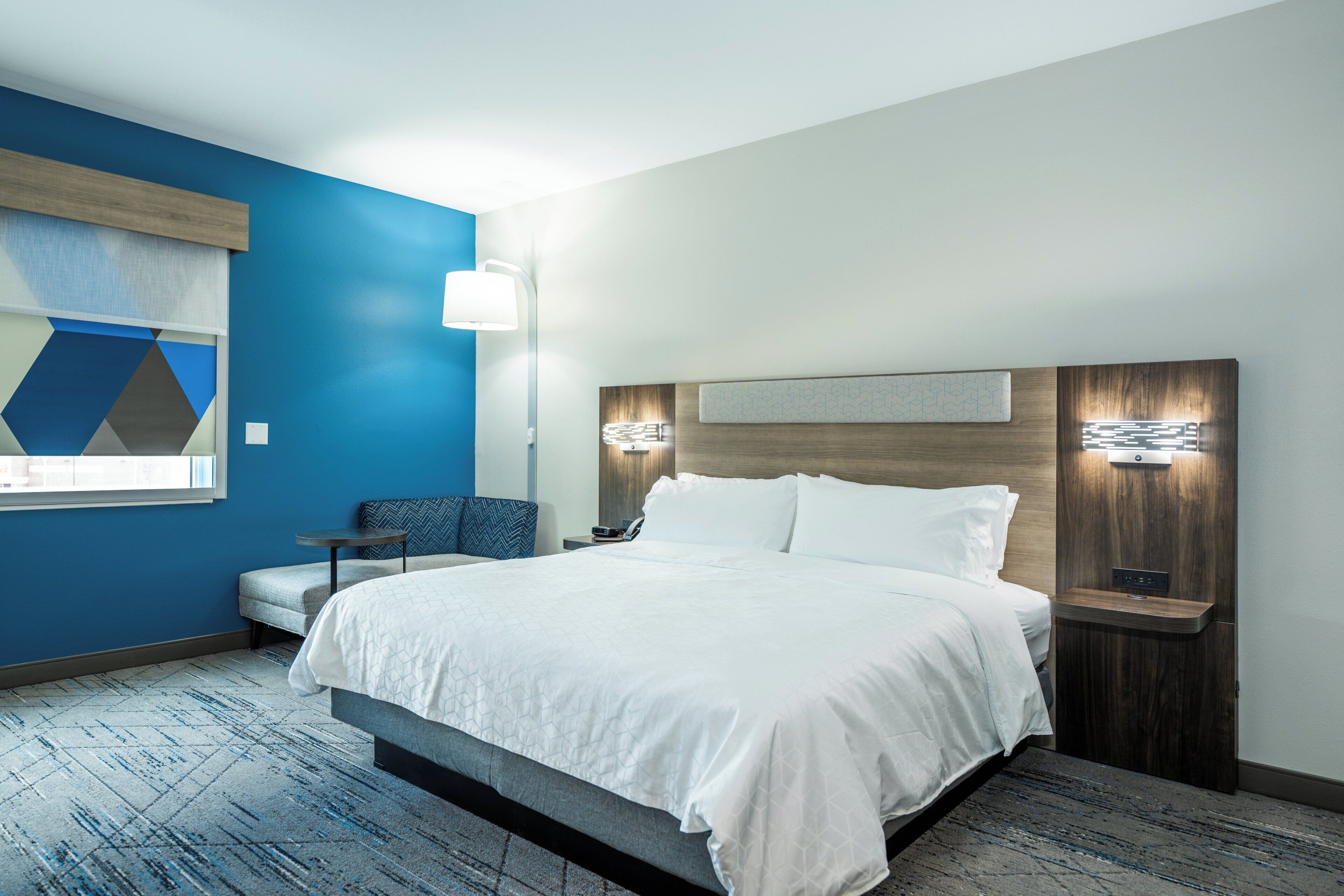 Quarto de hotel moderno com uma cama grande, uma mesinha de cabeceira, uma cadeira e uma luminária fixada na parede.