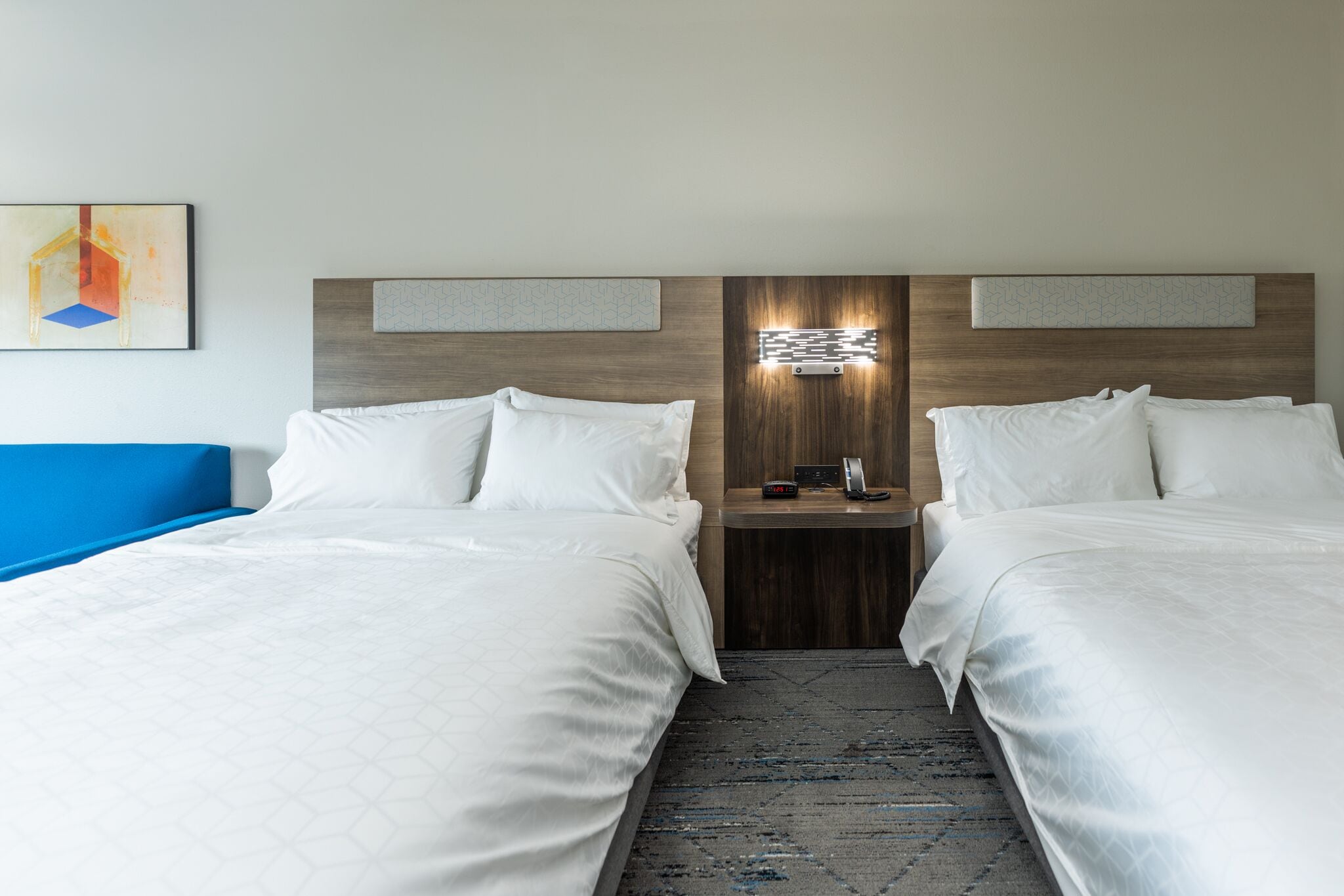 Quarto de hotel com duas camas, um sofá azul, uma mesa de cabeceira com telefone e despertador, e um candeeiro fixado na parede.