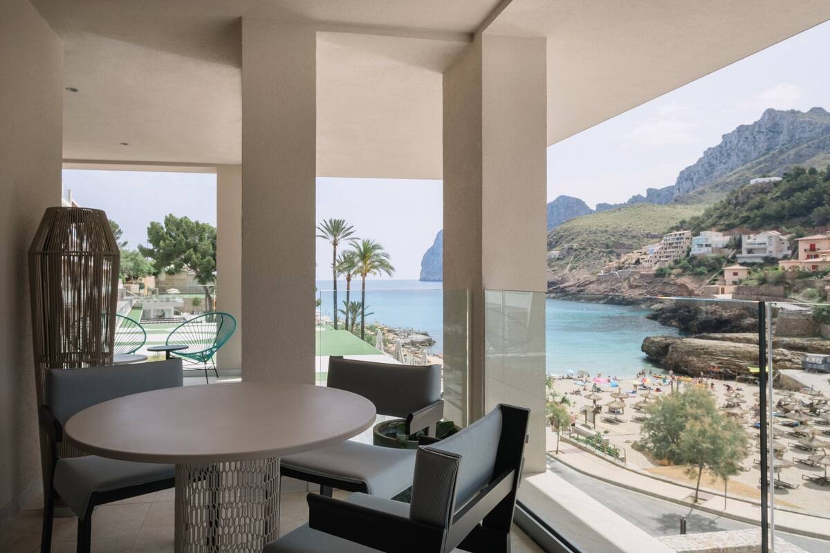Varanda com mesa e cadeiras, vista para uma praia com pessoas e edifícios.