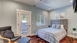 Quarto com uma cama grande, uma cadeira, uma mesinha com uma planta e duas janelas.