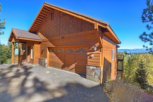 Uma cabana de madeira com garagem, varanda coberta e vista para a floresta e montanhas ao redor.