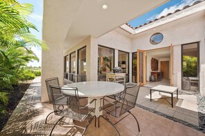Um pátio externo com uma mesa redonda e cadeiras, uma mesinha e vista do interior através de grandes portas de vidro.
