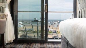 Quarto de hotel com cama, porta de vidro que dá para uma varanda com cadeiras e uma mesa, e vista para a praia.