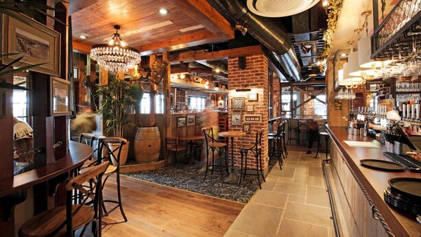 Um espaço de bar rústico com mesas de madeira, banquetas e parede de tijolos.