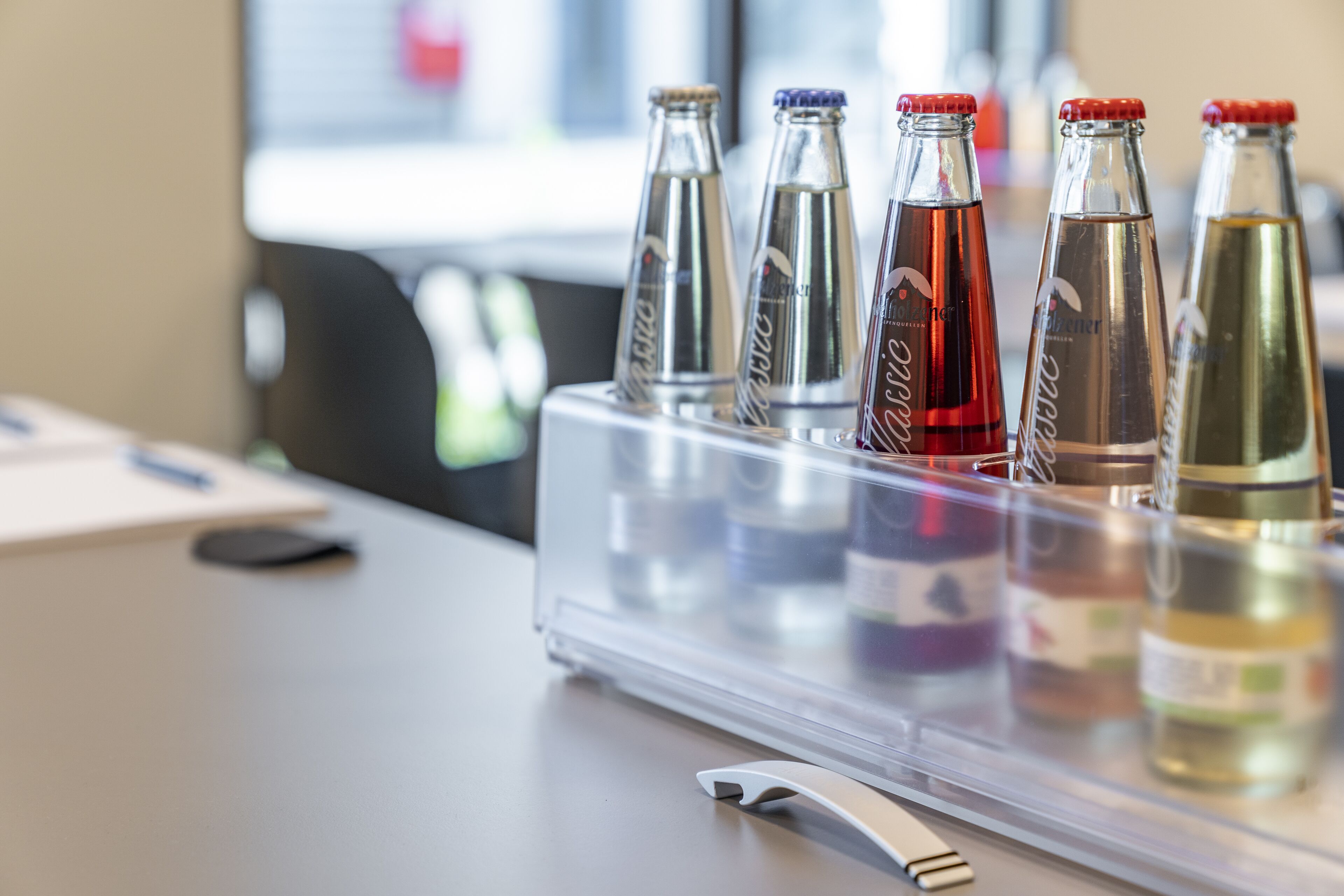Suporte de plástico transparente para garrafas com diversas garrafas, incluindo Coca-Cola, sobre uma mesa.