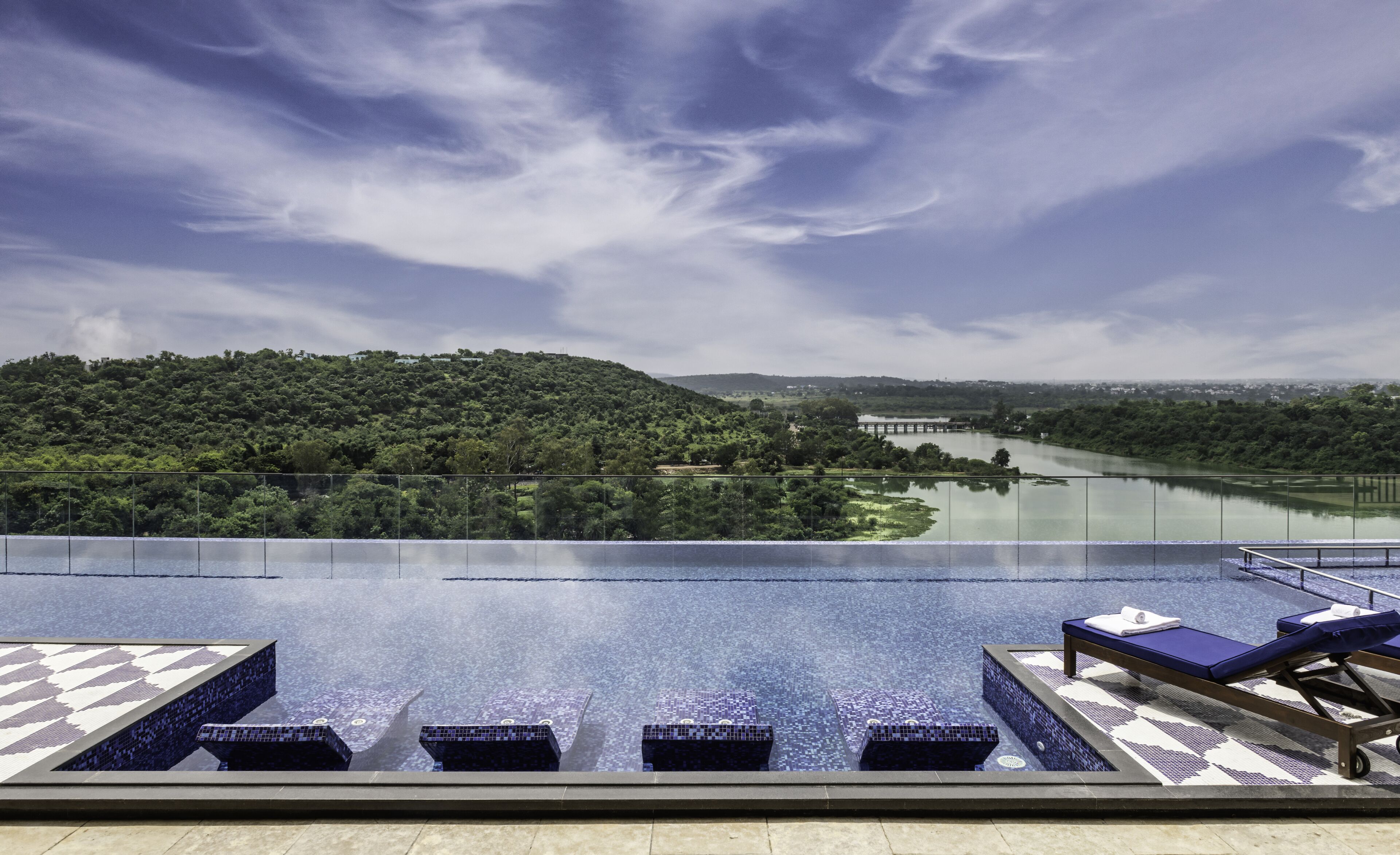 Piscina infinita com espreguiçadeiras e vista para um rio e colinas.