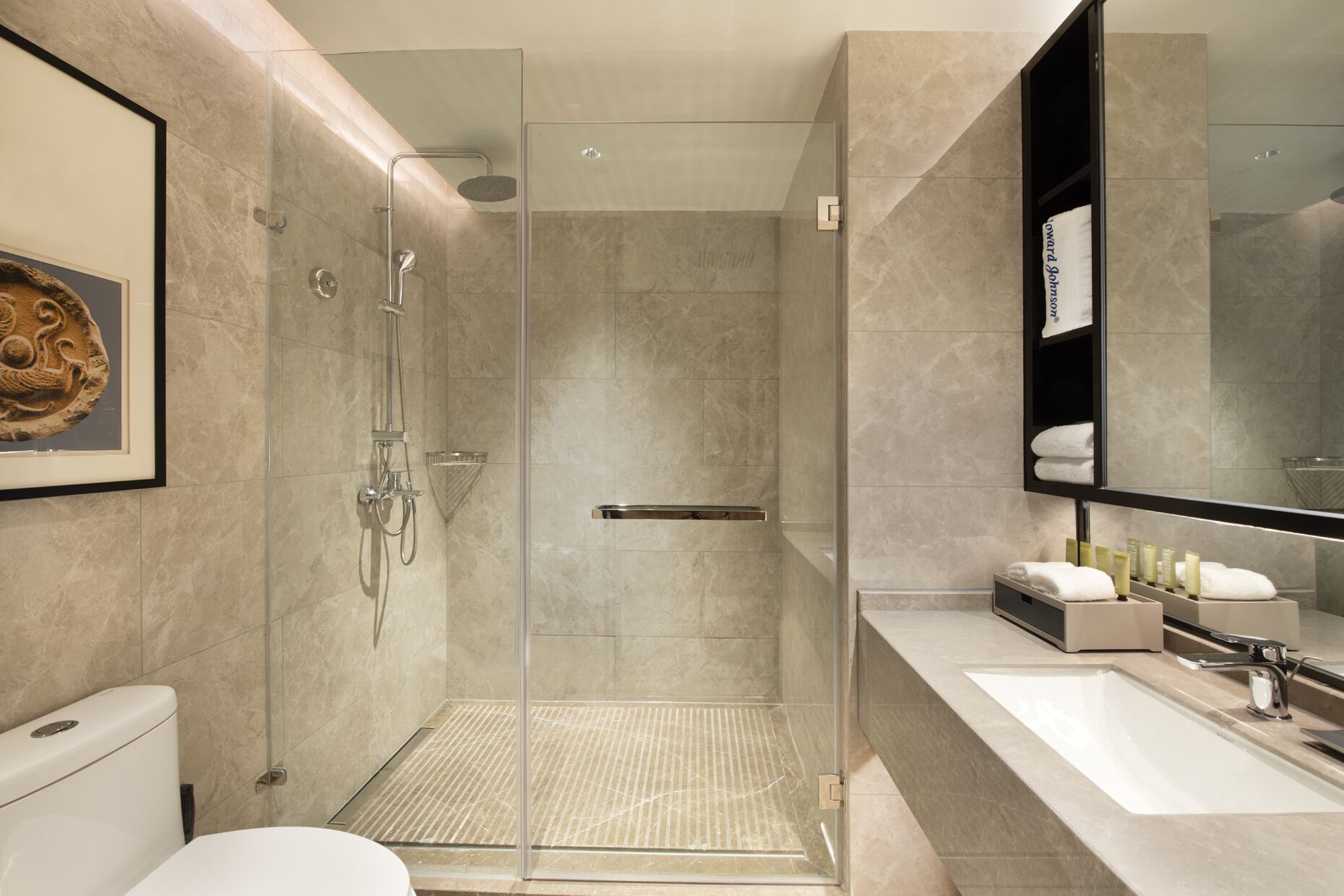 Um banheiro moderno com chuveiro envidraçado, vaso sanitário, pia com amenities e um quadro na parede.