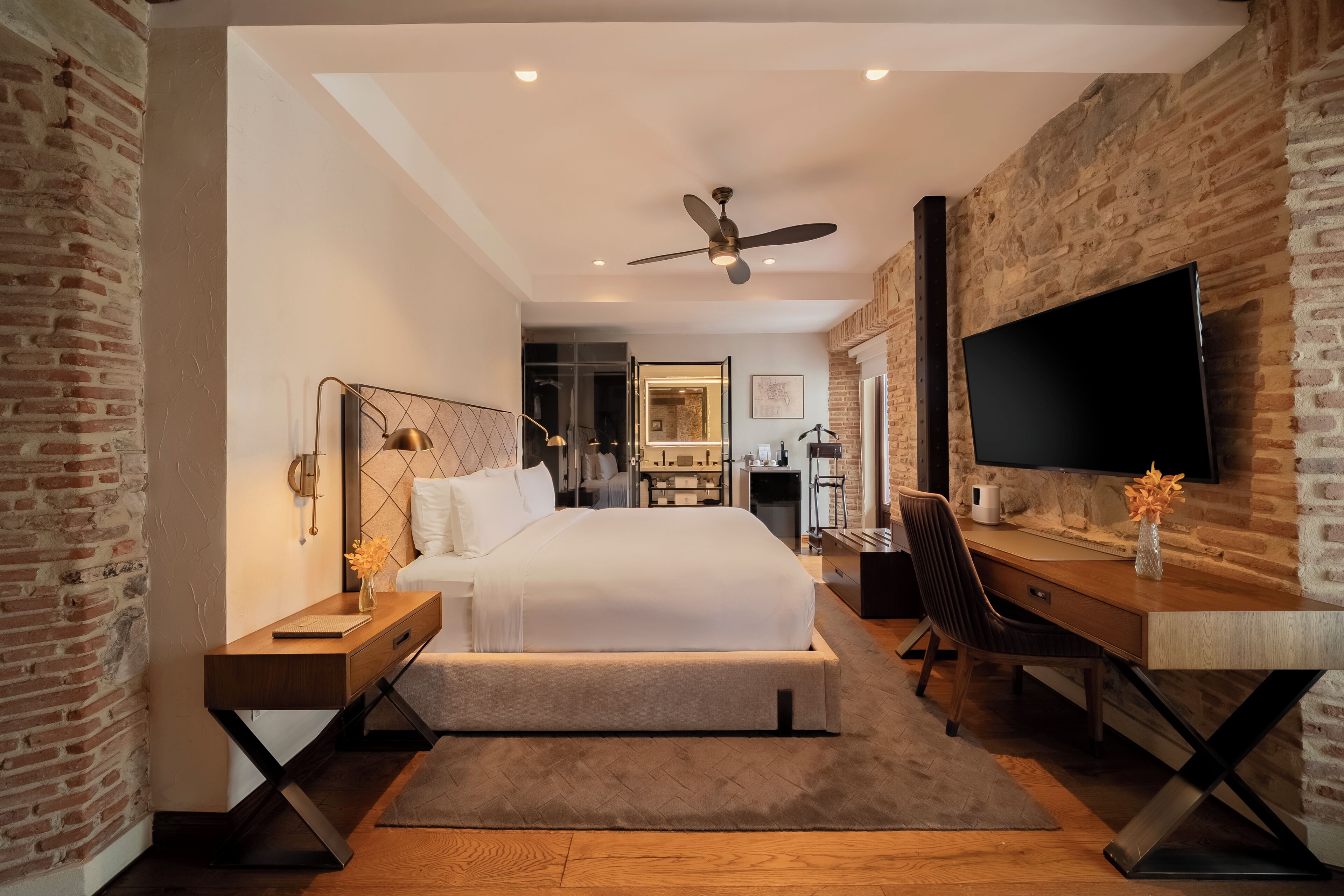 Quarto de hotel moderno com uma cama grande, uma escrivaninha, uma cadeira, uma televisão e um ventilador de teto.