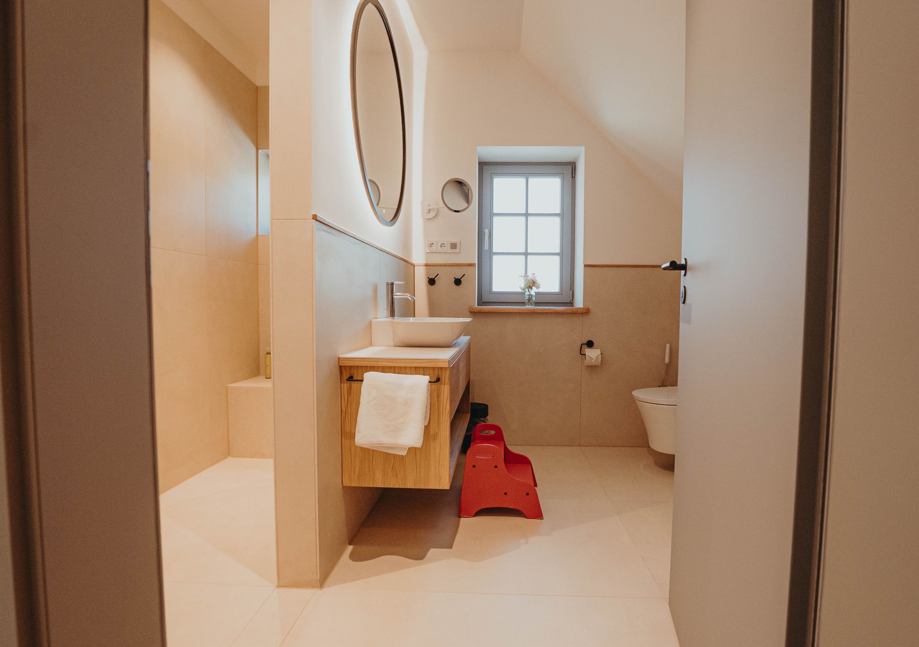 Uma casa de banho moderna com toucador de madeira, um banquinho vermelho, uma janela e uma sanita.