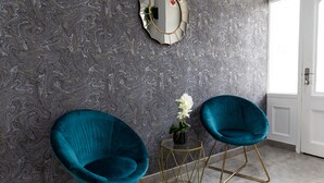 Um quarto com parede estampada em mármore, duas cadeiras cor turquesa, um espelho com moldura dourada e uma pequena mesa com um vaso de flores brancas.