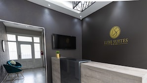 Uma área de recepção com parede escura, TV montada e logotipo da LUXE SUITES.