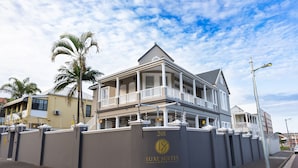 Um edifício de dois andares com varanda, com a inscrição "201 LUXE SUITES", cercado por um muro alto e palmeiras.