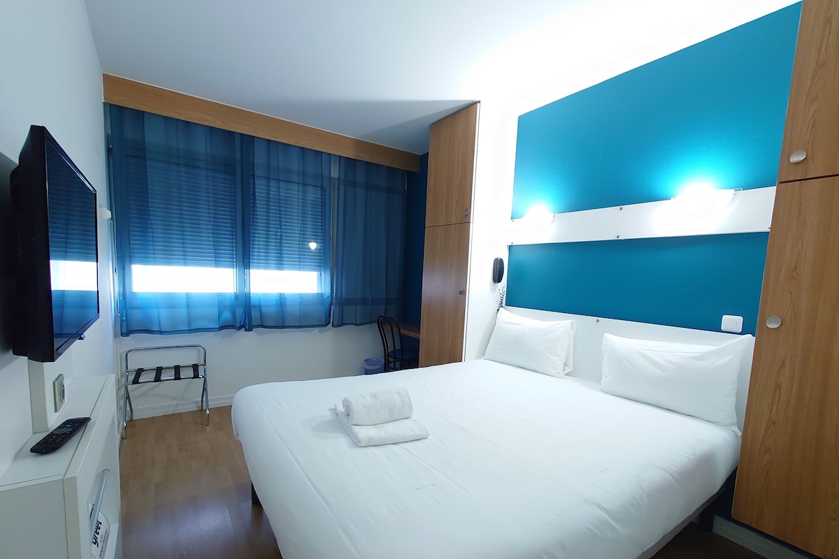 Quarto de hotel com uma cama grande, uma escrivaninha, uma cadeira, uma televisão e uma janela com cortinas azuis.