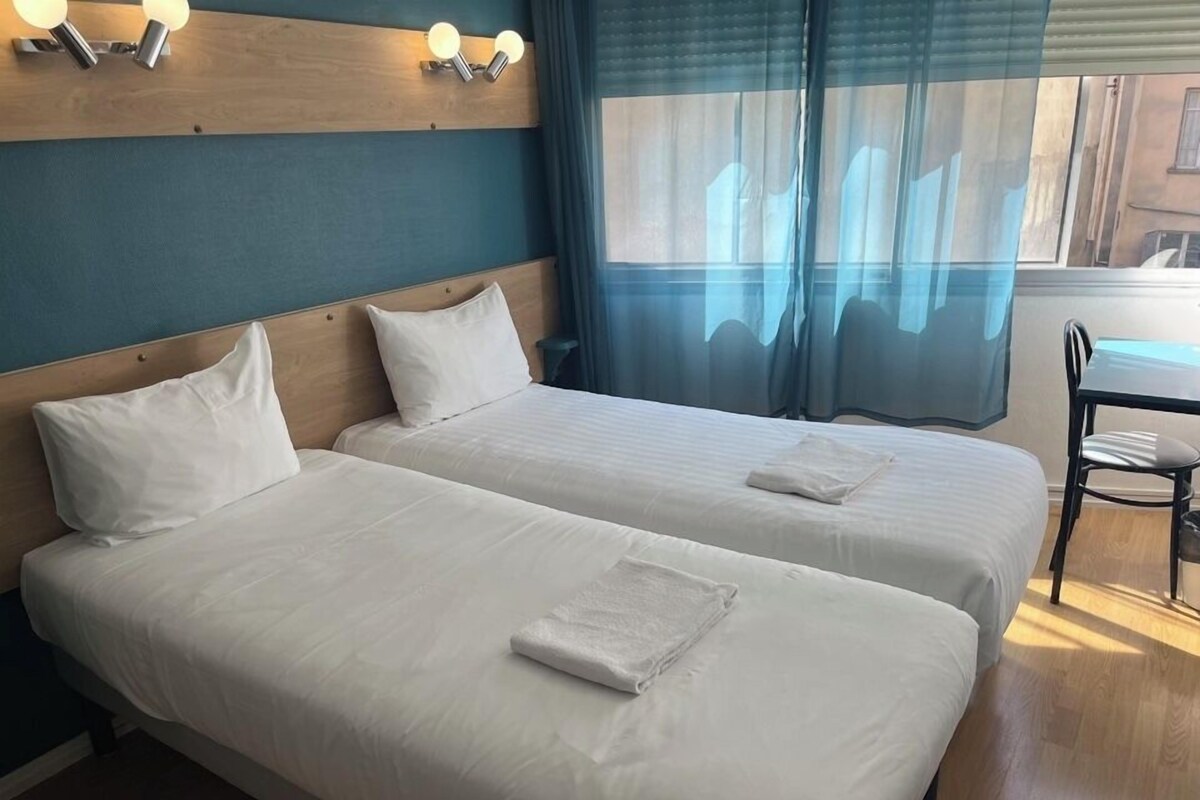 Quarto de hotel com duas camas, cabeceira de madeira, uma mesa e uma cadeira.
