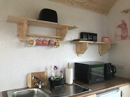 Cozinha com prateleiras de madeira contendo canecas e caixas, um micro-ondas e uma pia com tábua de corte.
