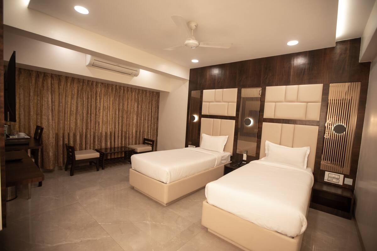 Quarto de hotel com duas camas, uma escrivaninha, uma cadeira e uma janela grande com cortinas.