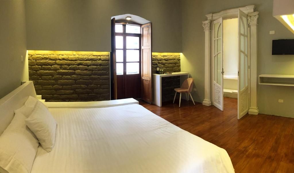 Quarto com uma cama grande, uma escrivaninha e uma cadeira. Apresenta uma parede de pedra e uma porta com painéis de vidro.