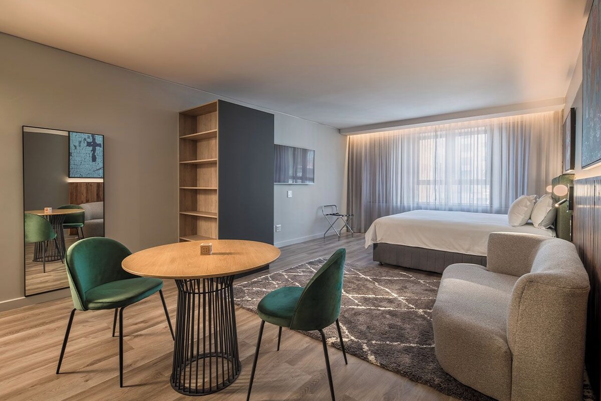 Quarto de hotel moderno com cama, sofá, mesa de jantar com cadeiras, espelho e estante.