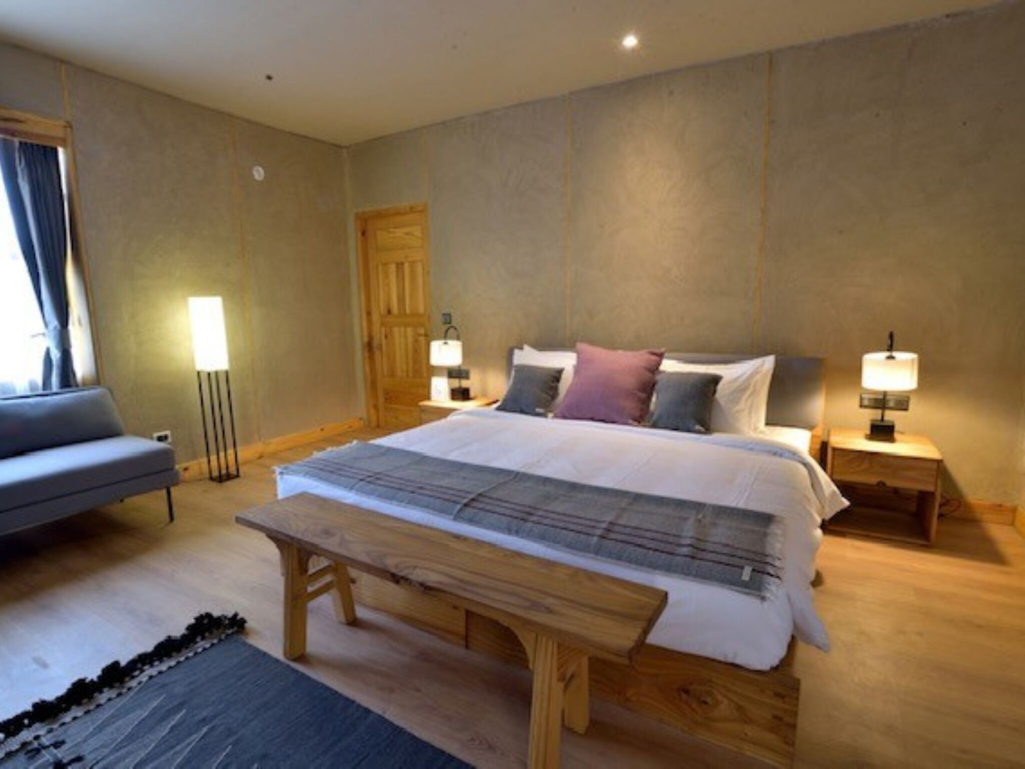 Quarto com uma cama grande, luminárias de cabeceira, um banquinho, um sofá e uma porta de madeira.