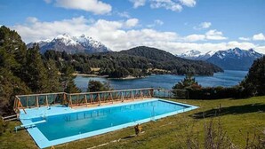 Piscina retangular com deck de madeira, cercada por gramado e montanhas ao fundo.