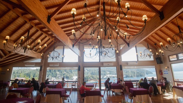 Restaurante com tetos de madeira alta e lustres, grandes janelas com vista para um lago e montanhas.