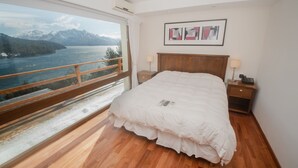 Quarto com uma grande janela que oferece vista para um lago e montanhas. Há uma cama com edredom branco, duas mesas de cabeceira e um quadro na parede.