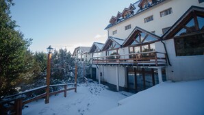 Área externa coberta de neve, com guarda-corpos de madeira e um edifício com grandes janelas e telhado inclinado.