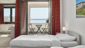 Quarto de hotel com cama, escrivaninha, duas cadeiras, mesa, janela com vista e um quadro na parede.