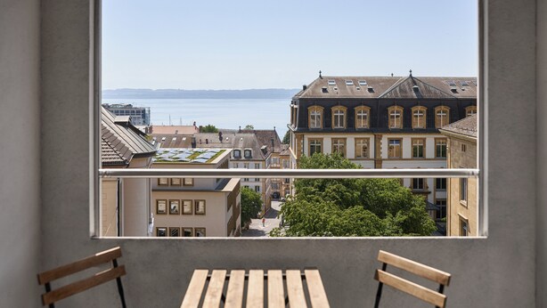 Vista da janela com duas cadeiras de madeira, mostrando um horizonte urbano com edifícios e um corpo d'água.