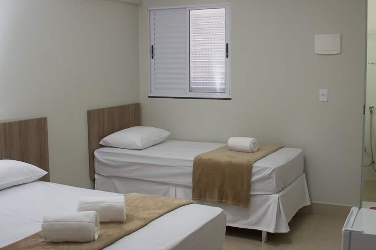 Quarto de hotel com duas camas, uma janela com persianas, um ar condicionado e um pequeno armário branco.