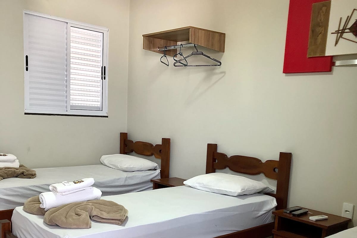 Quarto de hotel com duas camas de madeira, uma janela com persianas, um gancho para casacos na parede e uma pequena mesa de cabeceira de madeira.