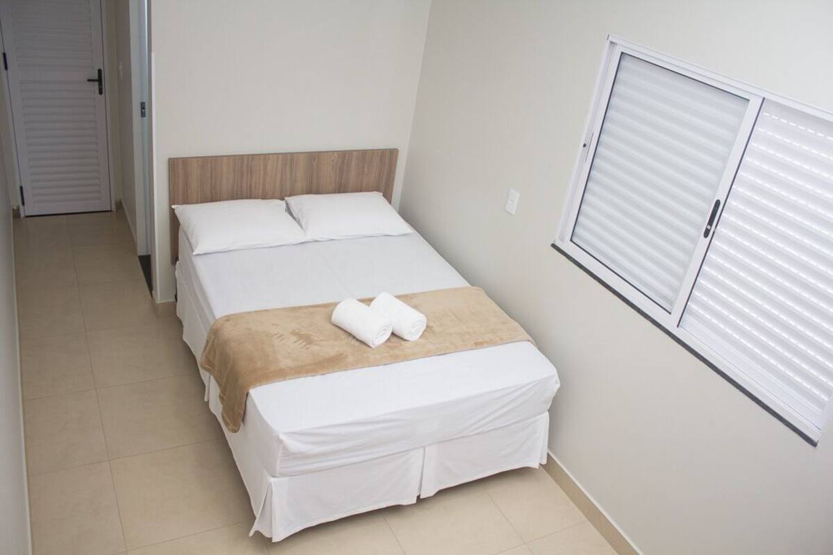 Uma cama de solteiro com cabeceira de madeira, roupa de cama branca e um cobertor bege. Há uma janela com persianas no lado direito.