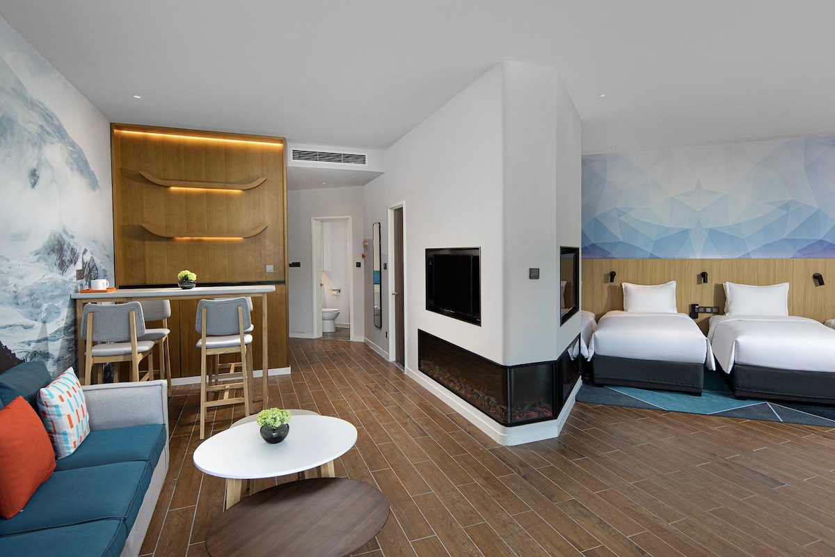 Quarto de hotel moderno com sofá, área de jantar com cadeiras, televisão na parede e lareira.