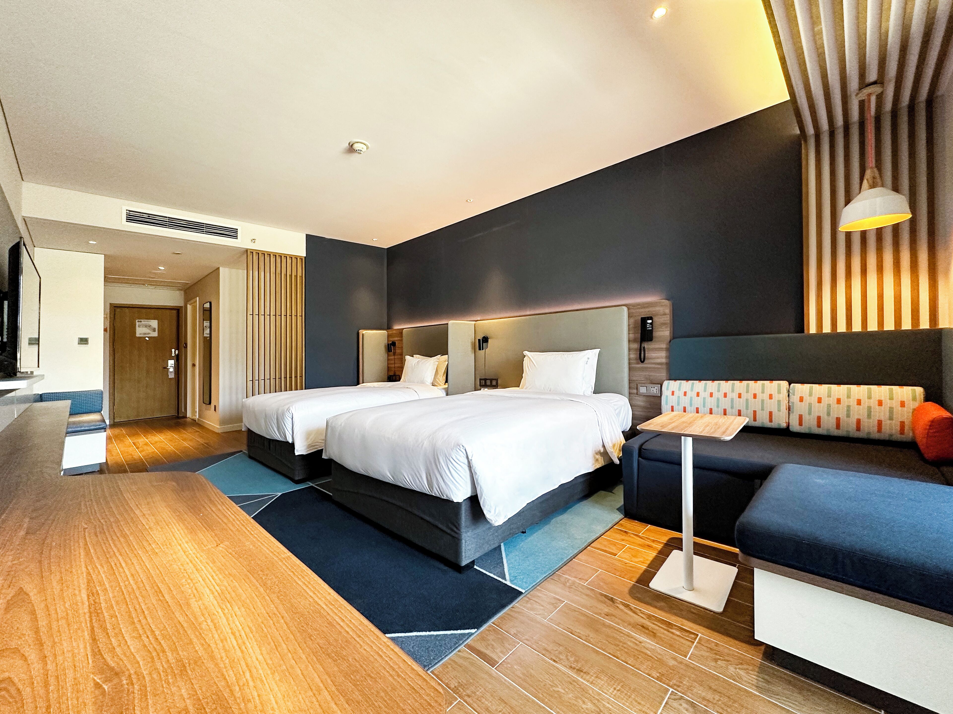 Quarto de hotel moderno com duas camas, um sofá, uma mesinha e um banheiro visível por uma porta aberta.