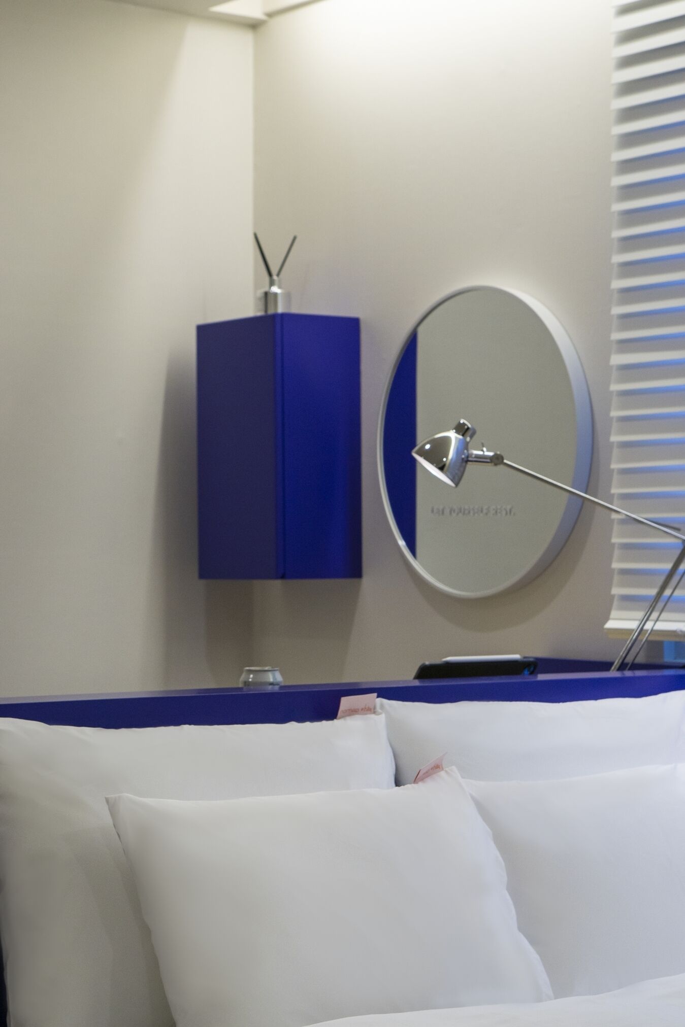 Quarto de hotel com cama, almofadas brancas, um armário azul, espelho redondo e abajur.