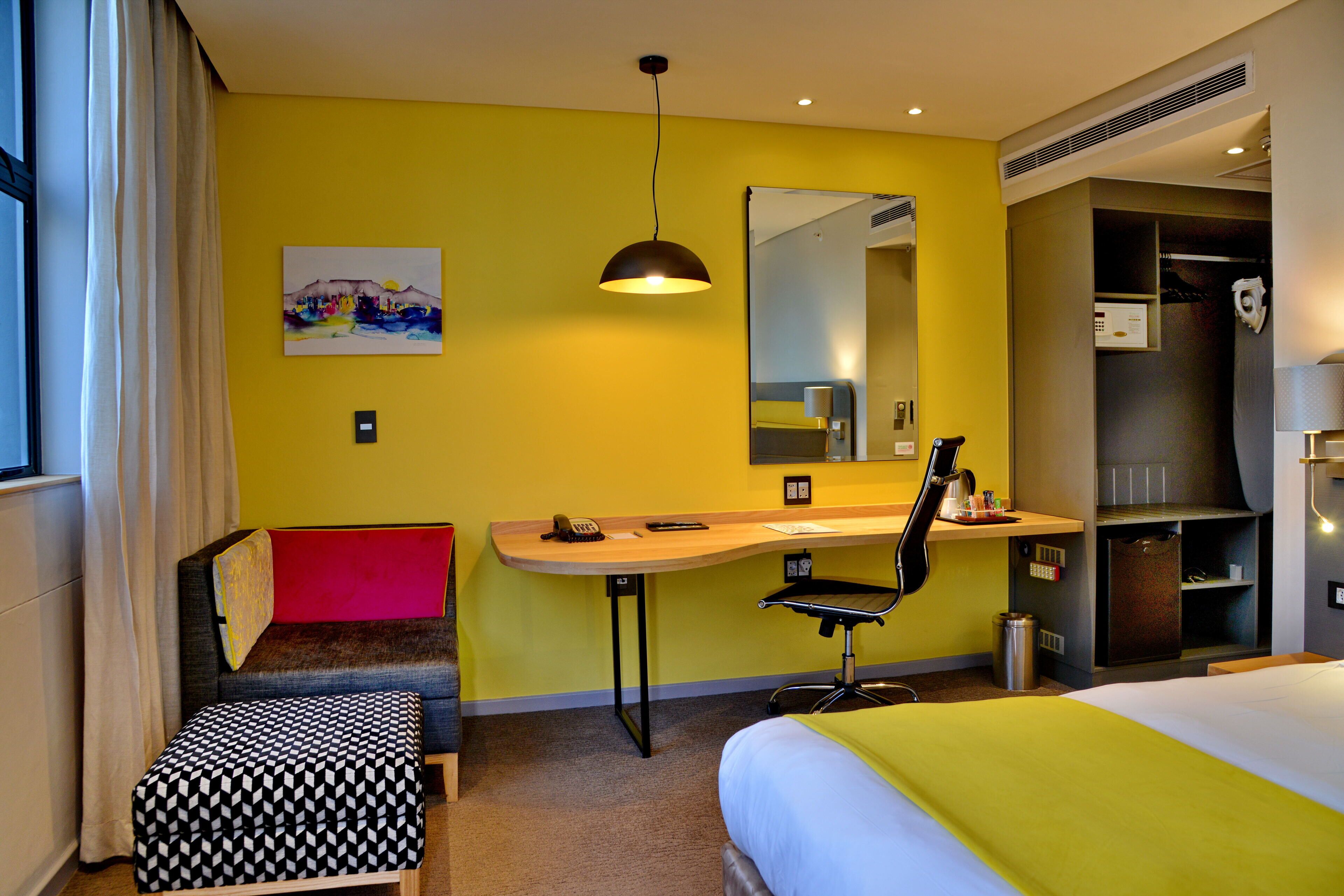 Quarto de hotel com cama, escrivaninha e cadeira, um pequeno sofá, espelho e janela com cortinas.