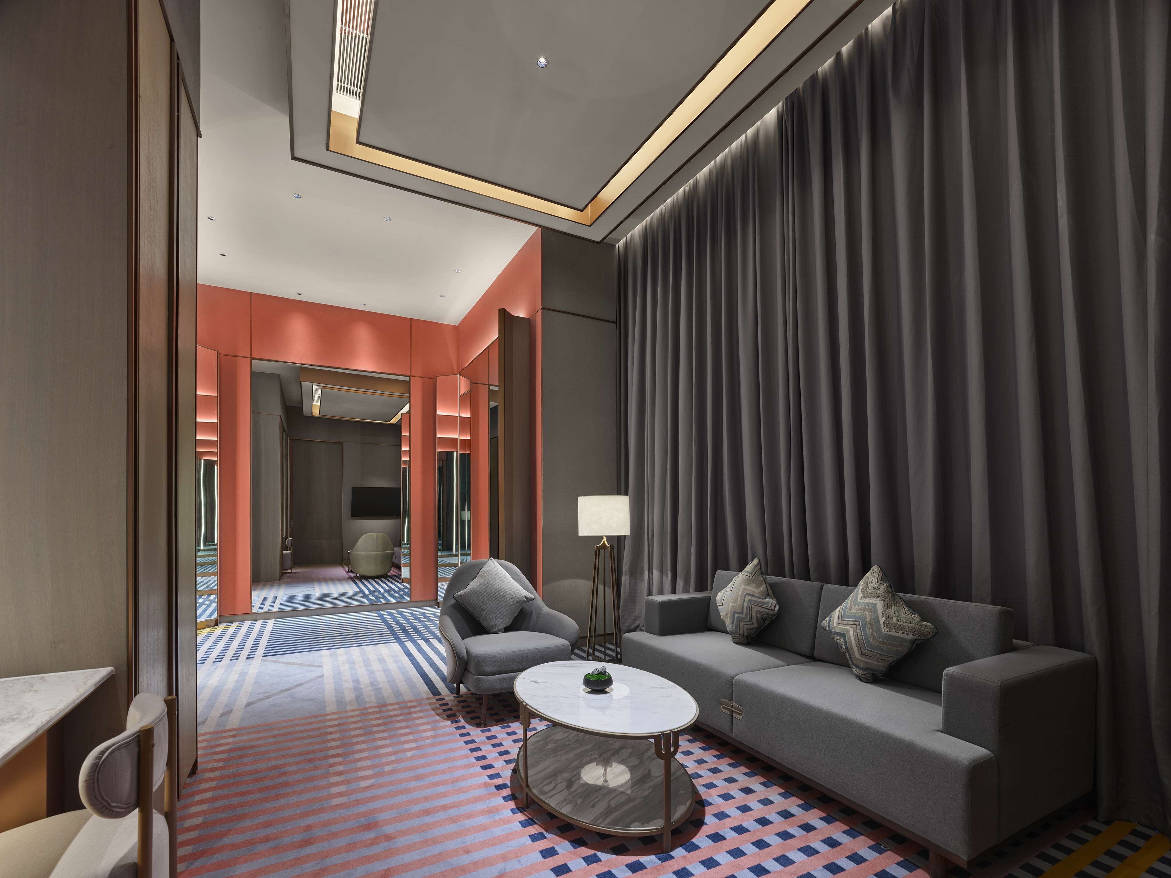 Quarto de hotel moderno com um sofá cinzento, poltronas, mesa de centro e um tapete estampado.