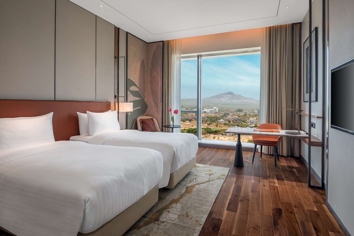 Quarto de hotel com duas camas, área de refeições e uma grande janela com vista para as montanhas.