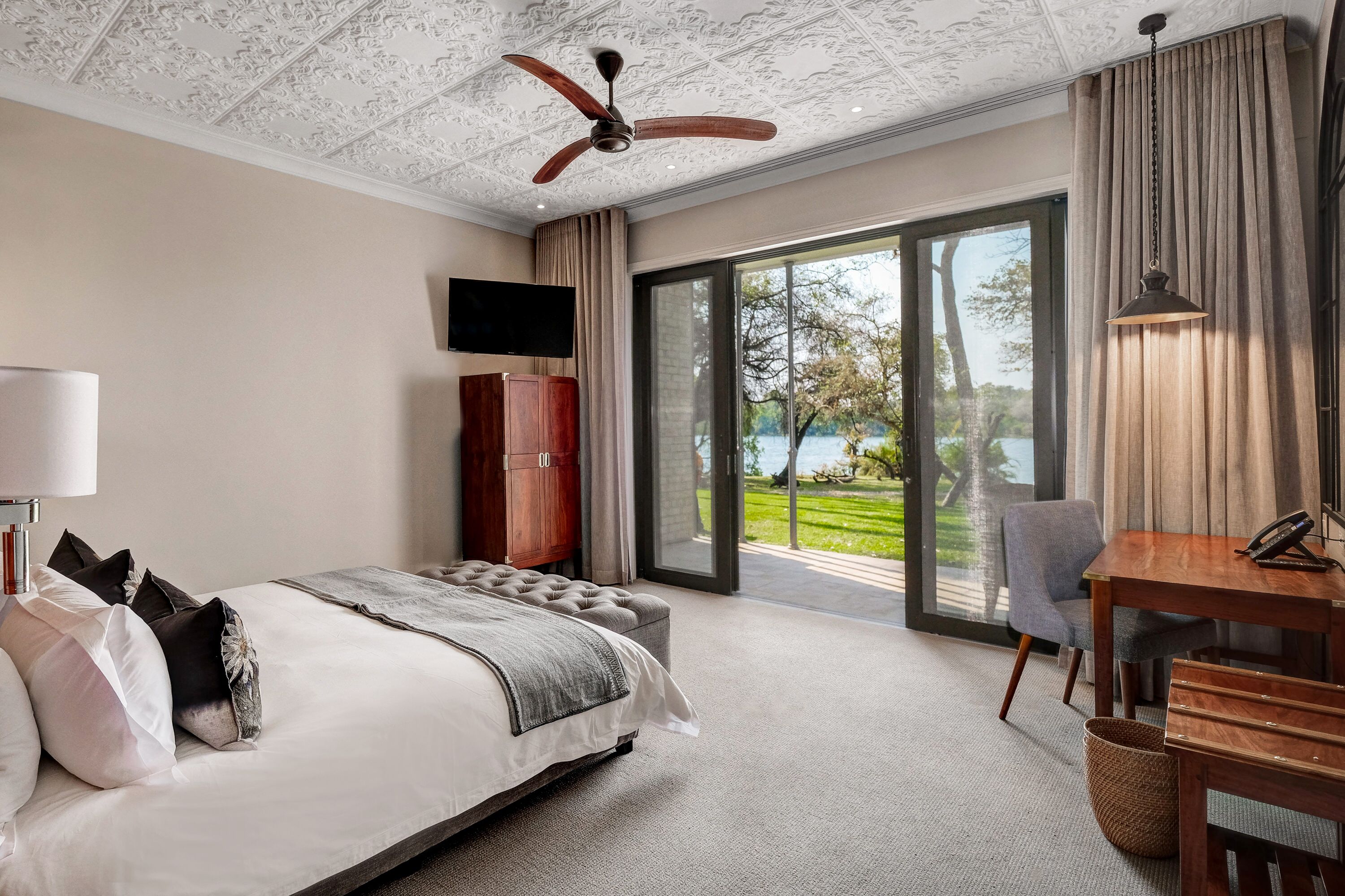 Quarto com cama, escrivaninha, cadeira, televisão e vista para o exterior.
