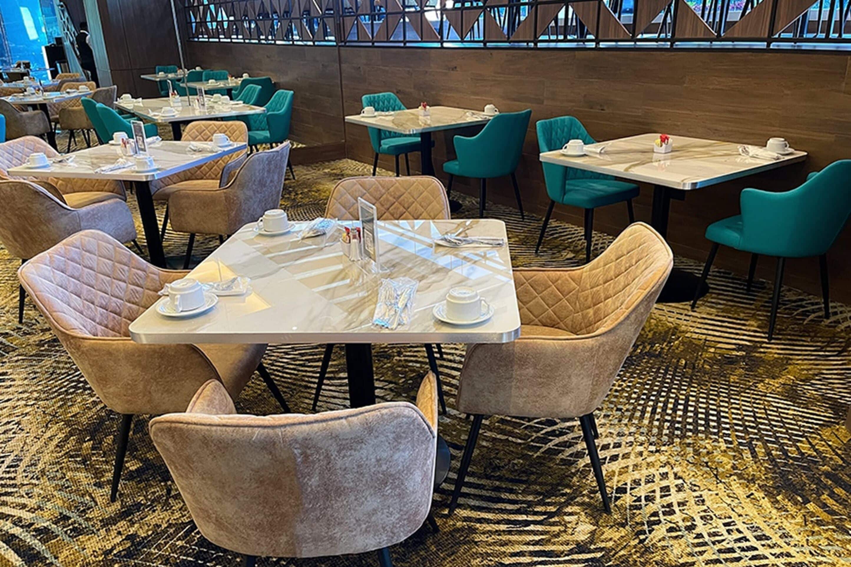 Um restaurante com mesas preparadas para refeições, com cadeiras acolchoadas e um tapete estampado.