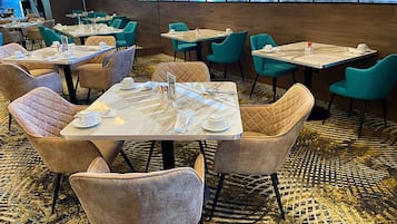 Um restaurante com mesas preparadas para refeições, com cadeiras acolchoadas e um tapete estampado.